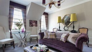 Uma sala de estar com um sofá roxo, um banquinho xadrez, uma mesa de centro de vidro e uma mesa de jantar com uma bandeja de itens de café da manhã.