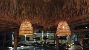 Área de piscina com cobertura de palha, espreguiçadeiras e um edifício moderno ao fundo.