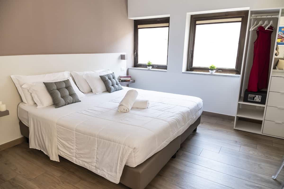 Uma cama bem arrumada com roupa de cama branca e várias almofadas, chão de madeira e um armário com roupas penduradas.