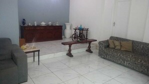 Uma sala de estar com um sofá cinza, um sofá estampado, um banco de madeira, uma mesinha com objetos decorativos e um armário em frente a uma parede azul.