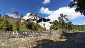 Uma casa de dois andares com um muro de pedra, um caminho de entrada pavimentado e vegetação exuberante, incluindo palmeiras.