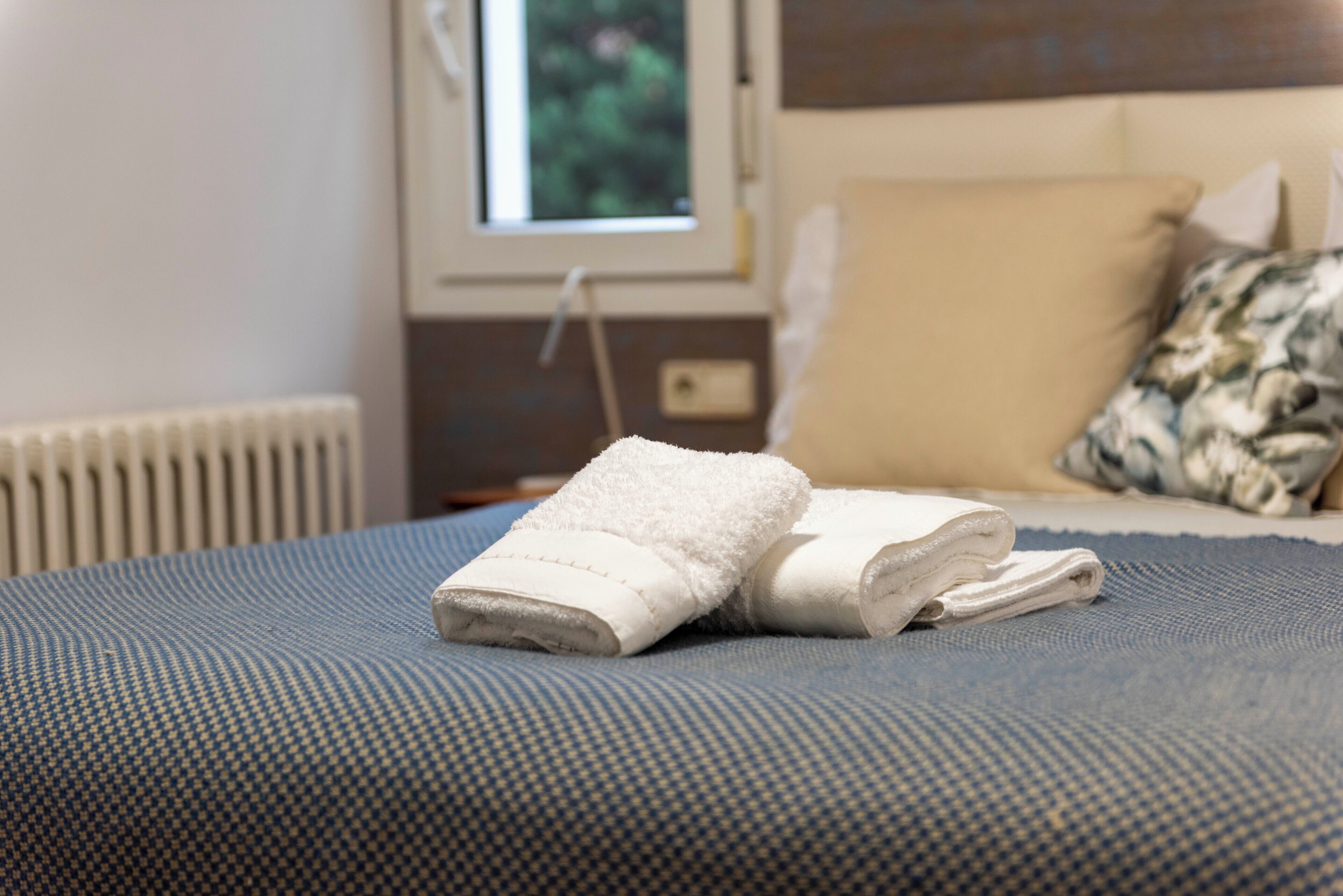 Cama bem arrumada com colcha azul estampada, duas toalhas brancas dobradas e uma janela com vista para o verde.
