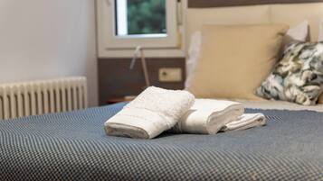 Cama bem arrumada com colcha azul estampada, duas toalhas brancas dobradas e uma janela com vista para o verde.
