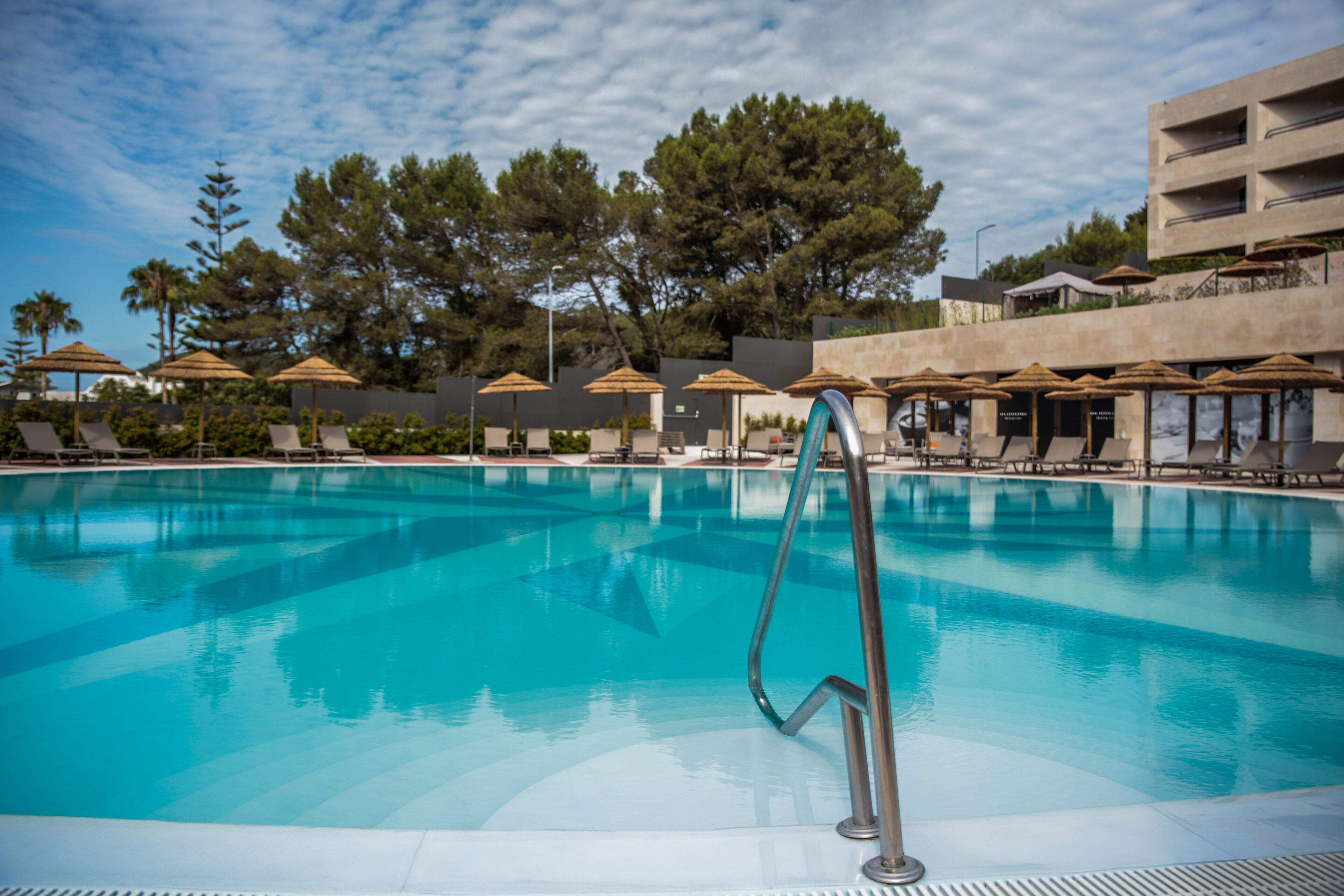 Uma piscina com espreguiçadeiras e guarda-sóis, cercada por árvores e um edifício.