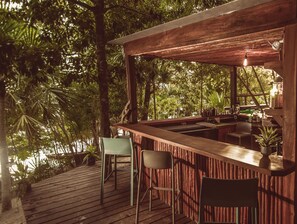 Um deck de madeira com área de bar, duas cadeiras e vista para vegetação exuberante.