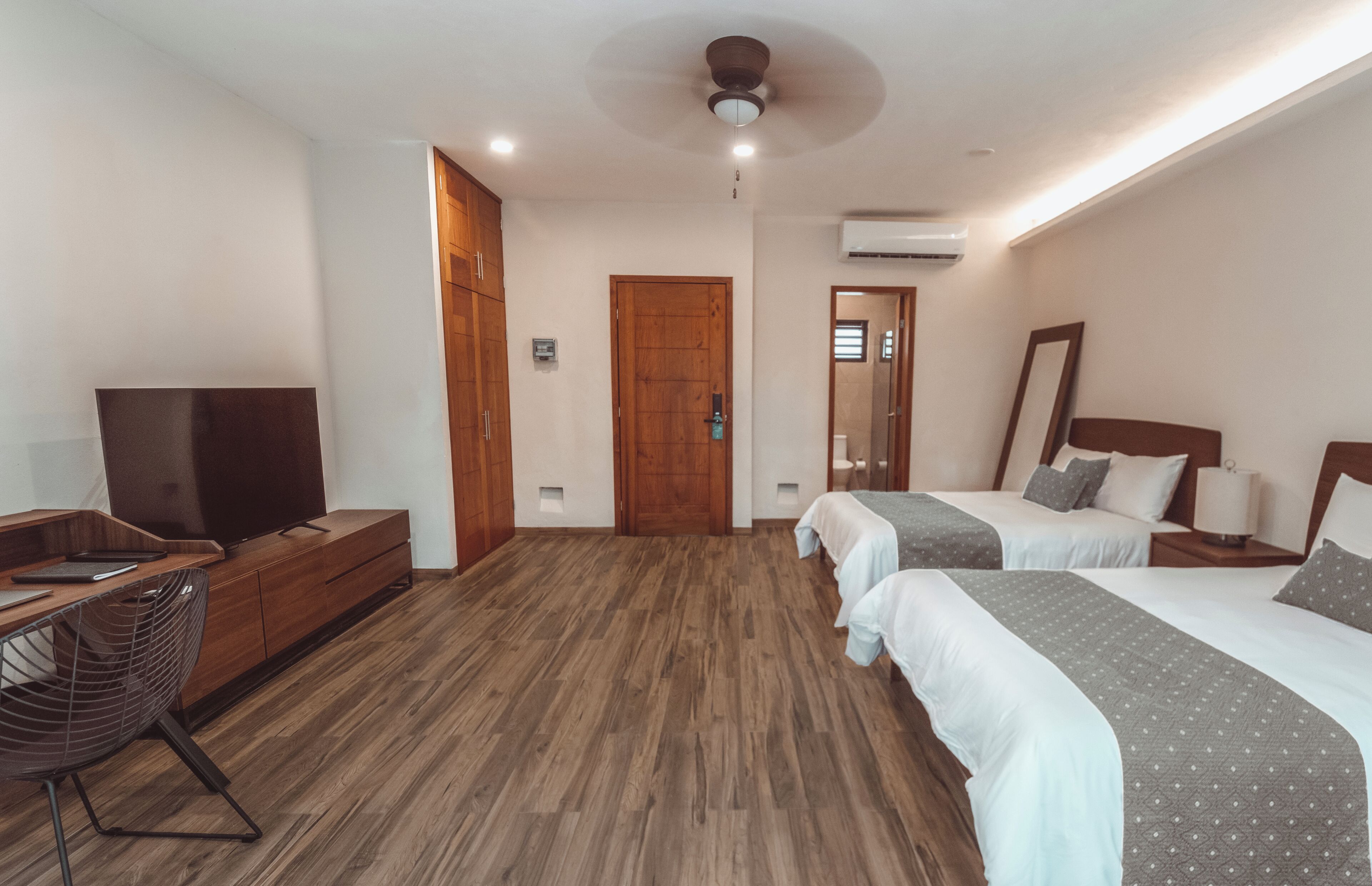 Quarto de hotel com duas camas, uma escrivaninha com cadeira, televisão e ventoinha no teto.