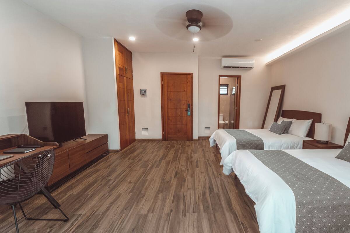 Quarto de hotel com duas camas, uma escrivaninha com cadeira, televisão e ventoinha no teto.