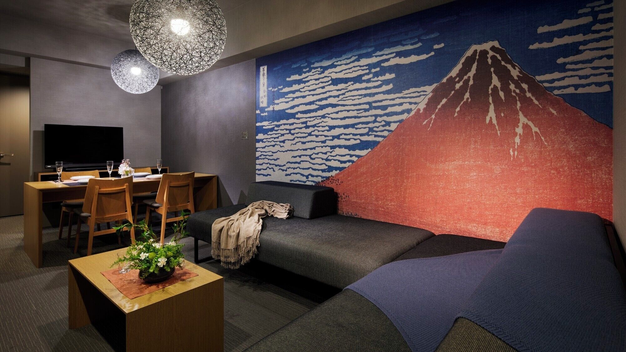 Quarto de hotel moderno com um grande mural na parede retratando o Monte Fuji, um sofá, uma mesa de centro com uma planta, uma escrivaninha com uma TV de tela plana e duas luminárias pendentes esféricas.