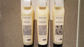 Três frascos de produtos Pérceige: shampoo, condicionador e gel de banho, dispostos sobre uma superfície azulejada.