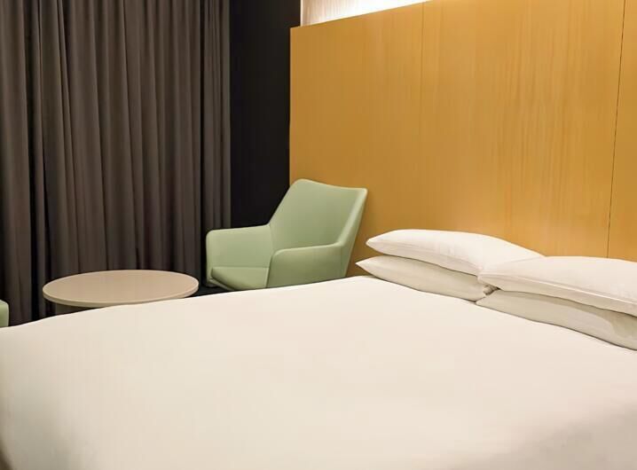 Quarto de hotel com cama, uma cadeira verde, uma mesinha redonda e cortina.