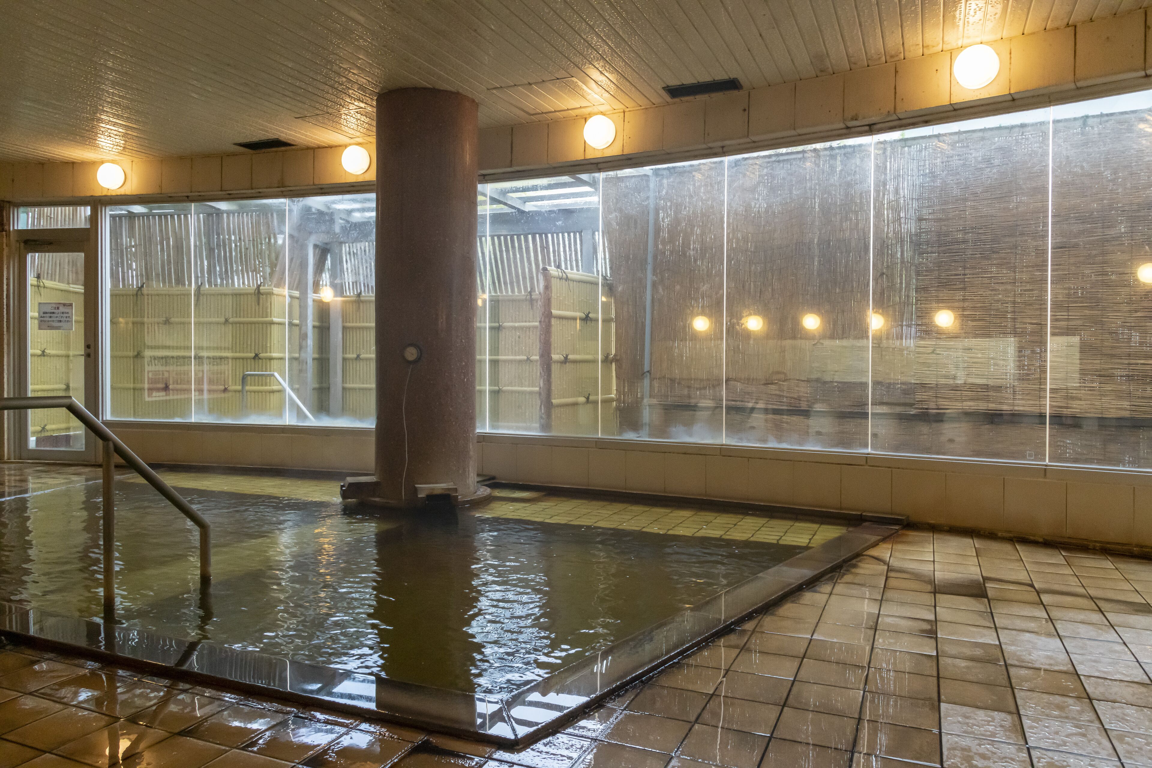 Uma piscina ampla em ambiente interno, com paredes de vidro transparente, piso revestido e um pilar central.