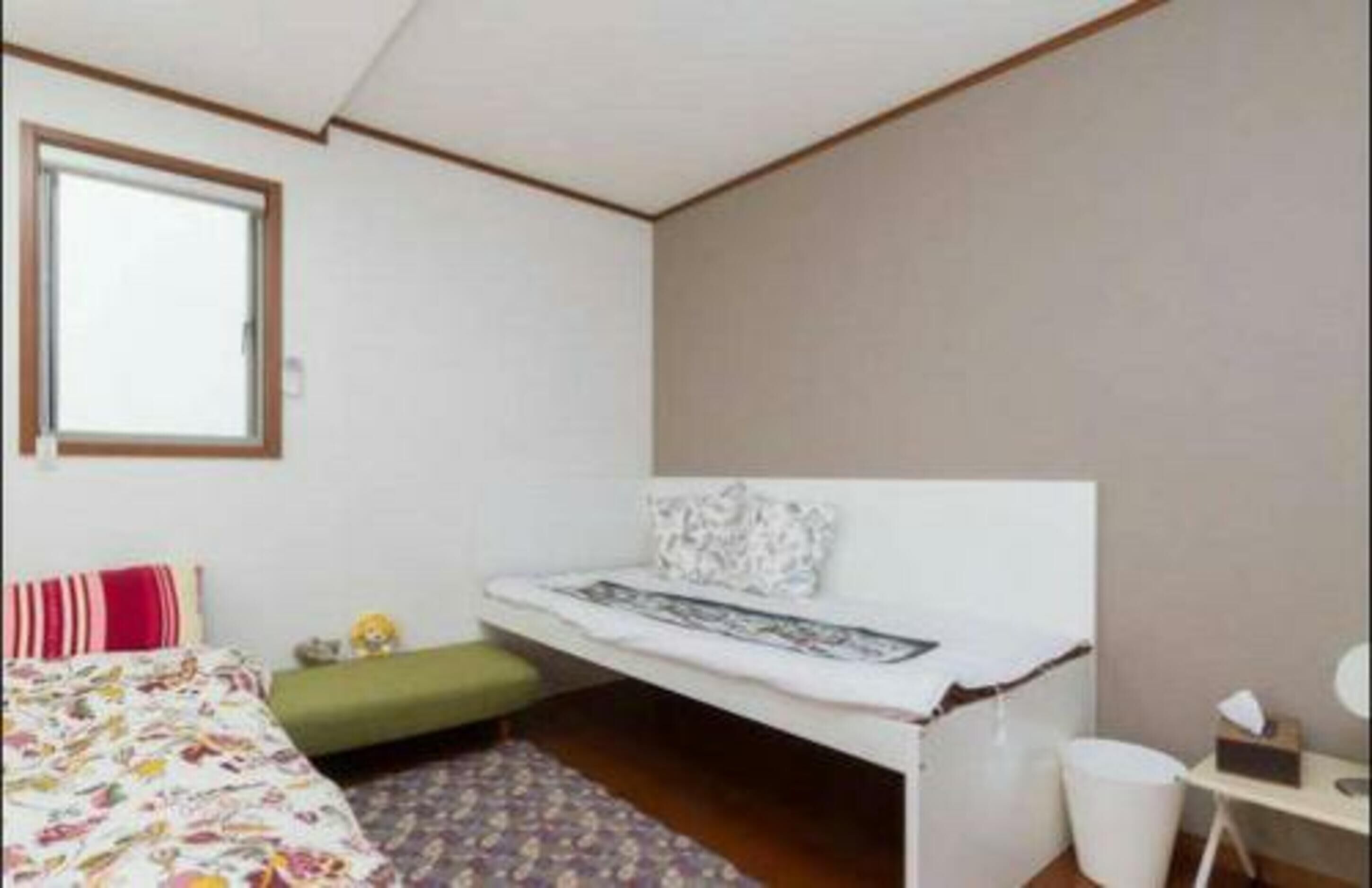Quarto pequeno com uma cama de solteiro, janela, mesa de cabeceira, um banquinho verde e uma pequena lixeira.