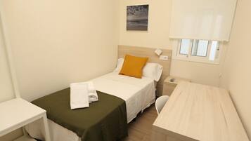 Um quarto pequeno e bem arrumado, com uma cama, mesa de cabeceira, cadeira e escrivaninha. Há uma janela com persianas e um quadro na parede.