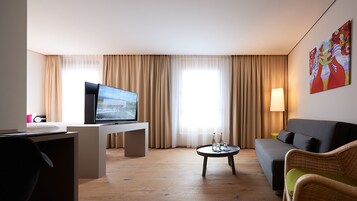 Quarto de hotel moderno com televisão de tela plana em uma escrivaninha, um sofá, uma pequena mesa redonda e uma janela grande com cortinas.