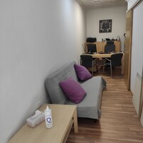 Quarto compacto com um sofá cinza, duas almofadas roxas, uma mesa de madeira com uma garrafa e uma caixa, e uma escrivaninha com um computador e uma cadeira ao fundo.