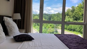 Quarto de hotel com uma cama grande, uma mesa de cabeceira, um abajur e uma janela com vista panorâmica.