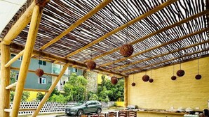 Uma área de refeições ao ar livre com estruturas de bambu, toalhas de mesa quadriculadas e decorações esféricas penduradas.