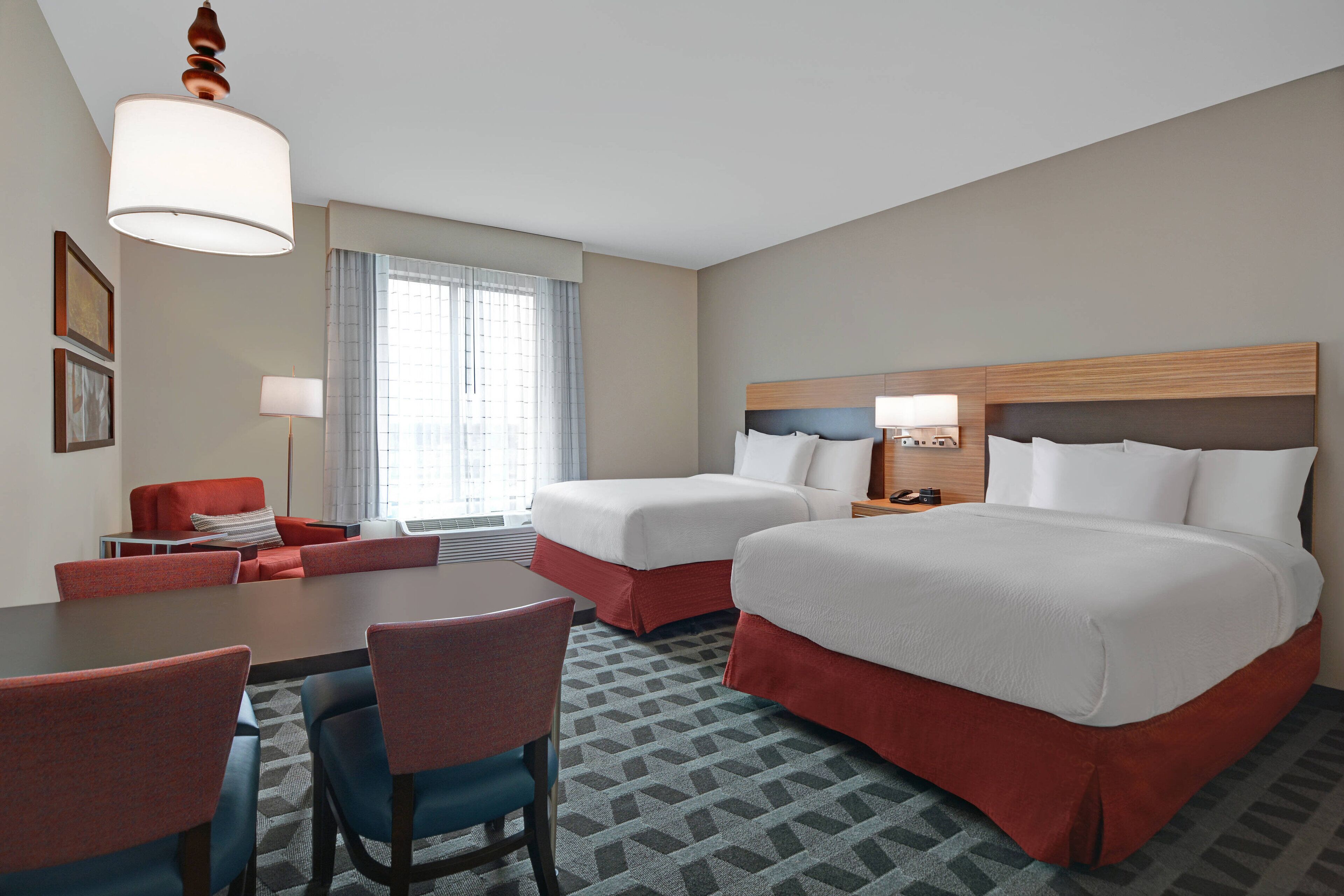 Quarto de hotel com duas camas, uma mesa de jantar com cadeiras, um abajur e uma janela com cortinas.