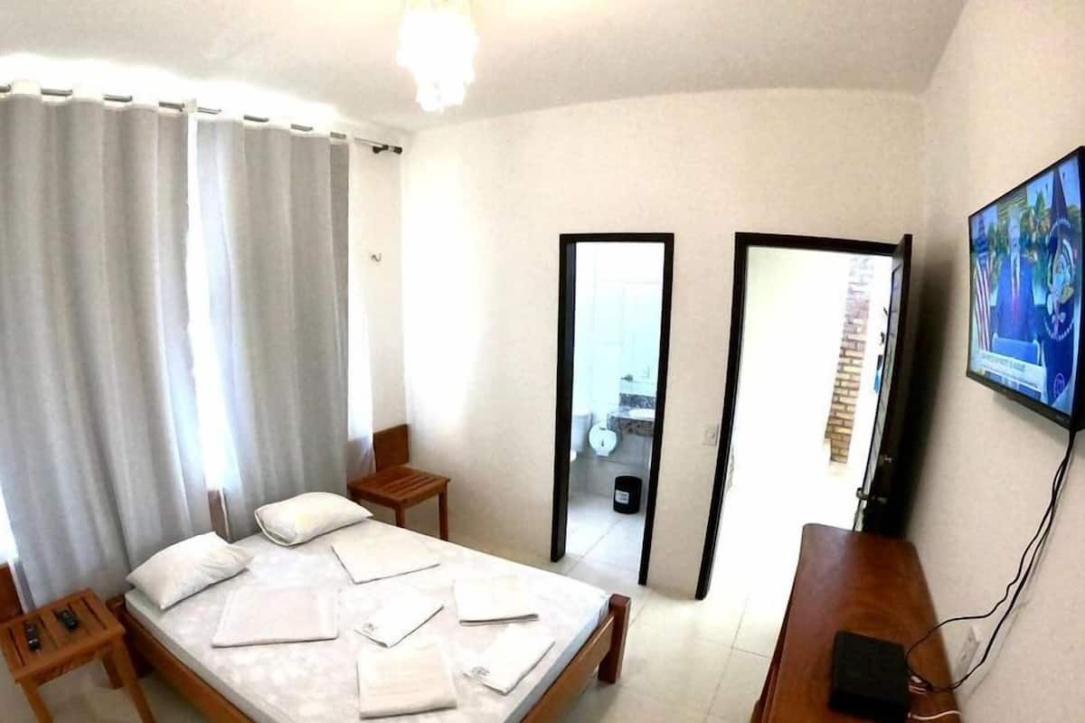 Quarto de hotel com cama, escrivaninha, televisão e banheiro visível através de uma porta aberta.