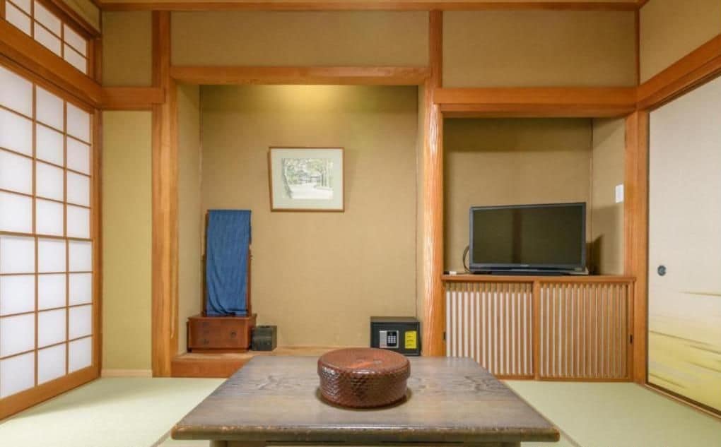 Um quarto japonês tradicional com tatames, uma mesa baixa com uma cesta de palha, uma televisão sobre um suporte de madeira e uma porta deslizante.