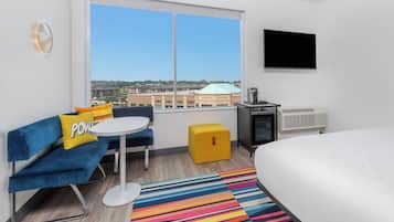 Quarto de hotel com uma janela grande, uma cama, uma mesa pequena, um sofá azul, um tapete colorido listrado e uma televisão de tela plana.