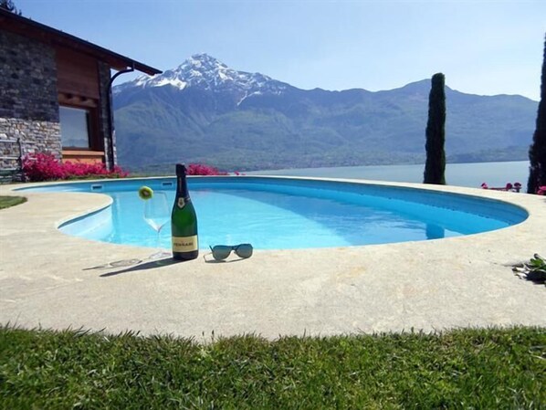Piscina com uma garrafa de champanhe e óculos de sol na borda, cercada por um edifício de pedra e vista para a montanha.