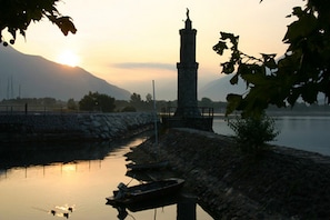 Um rio com um muro de pedra e uma torre, barcos atracados, e uma montanha ao fundo.