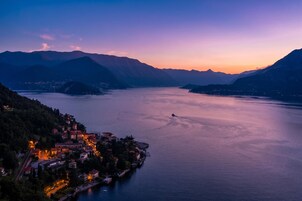 Uma cidade à beira de um lago, com edifícios ao longo da margem, um barco na água e montanhas ao fundo.