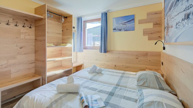Quarto de hotel moderno com paredes de madeira, uma cama grande com roupa de cama listrada, uma janela com cortinas azuis e um quadro de uma paisagem nevada.