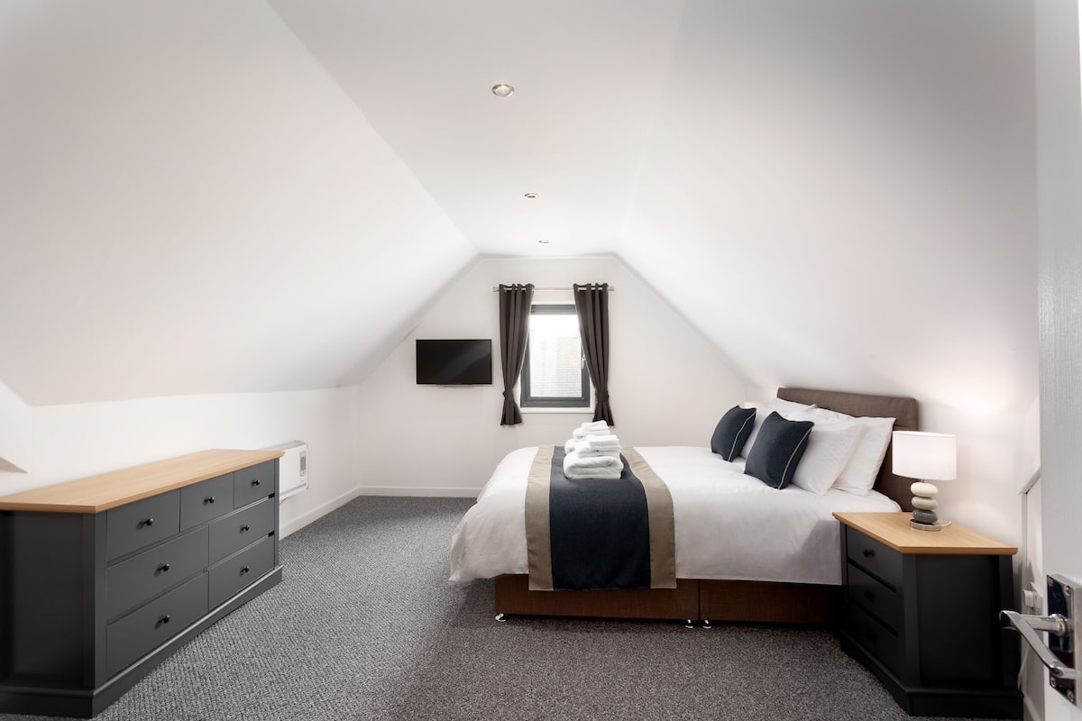 Quarto de hotel moderno com uma cama bem arrumada, cômoda e mesa de cabeceira com abajur.