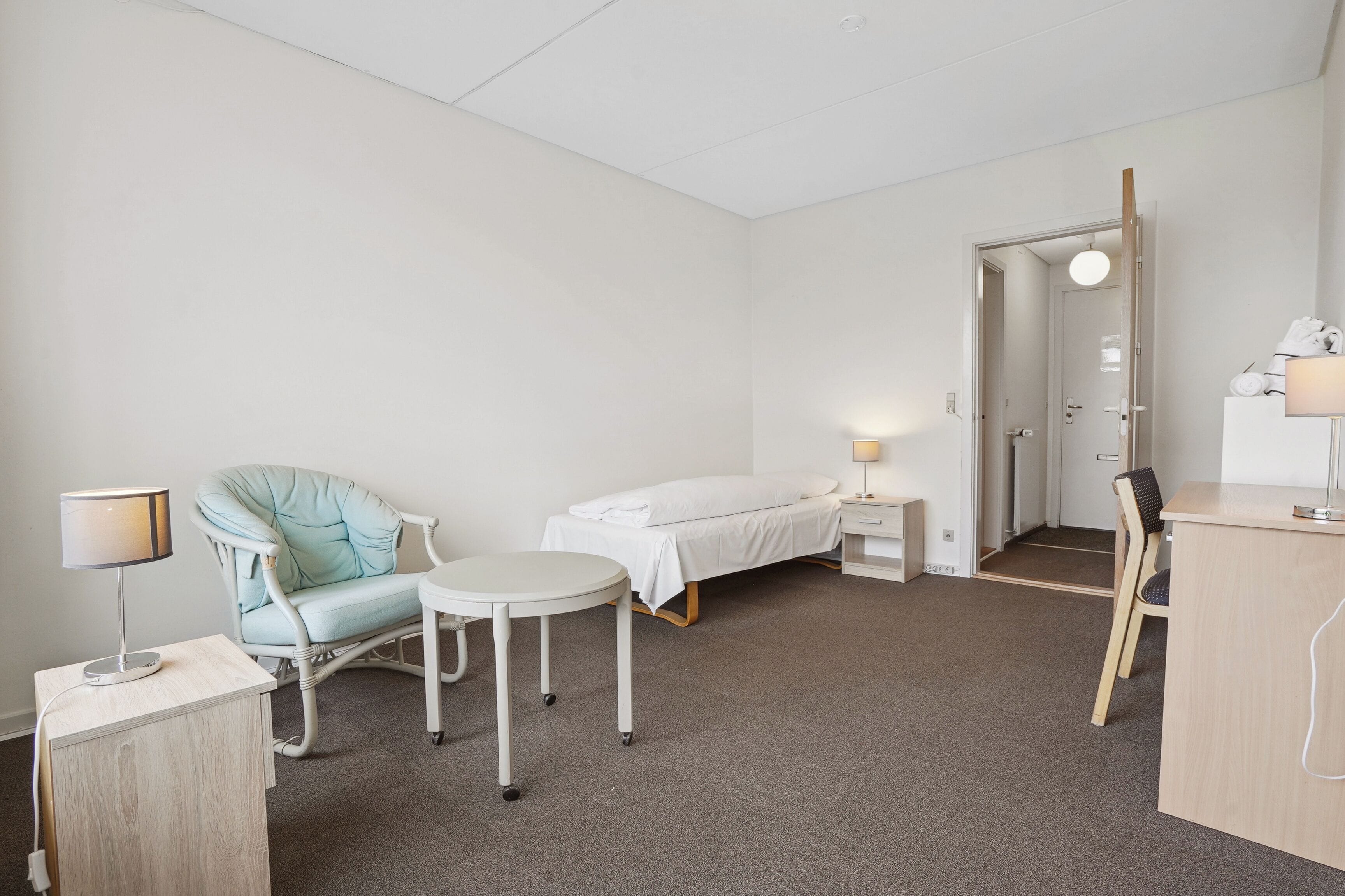 Quarto de hotel com cama, uma cadeira, escrivaninha e luminária.