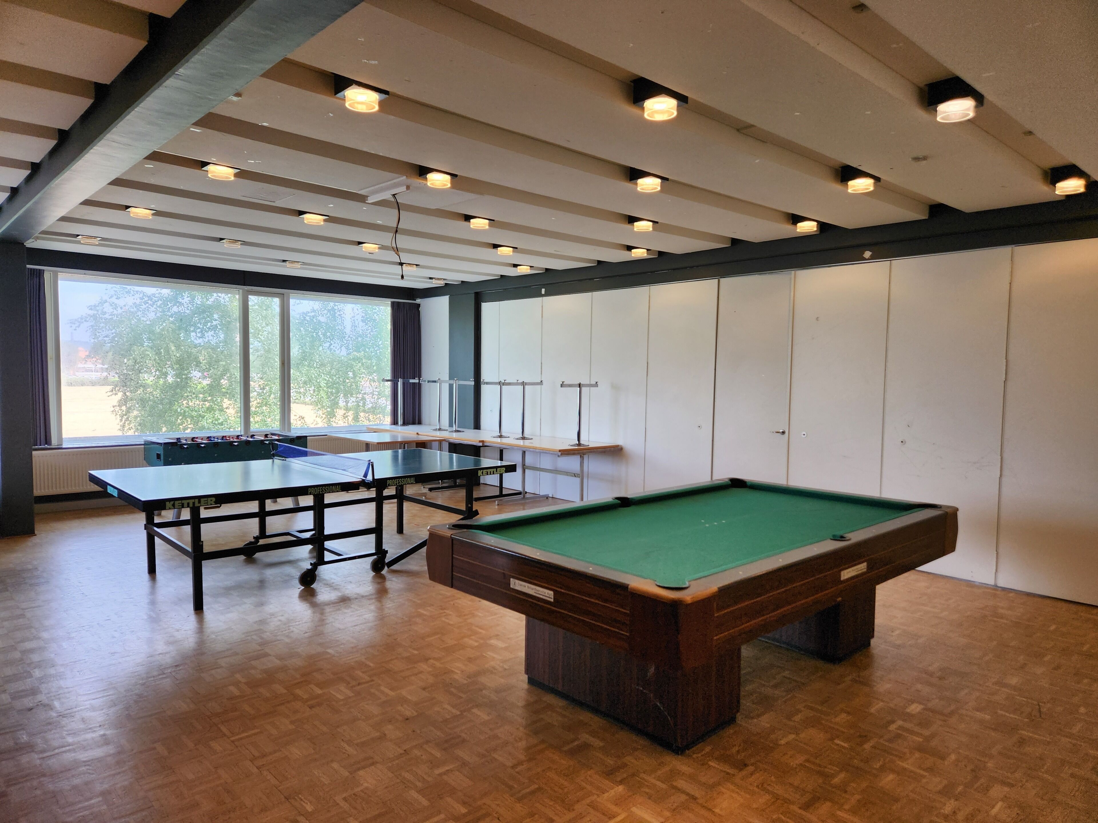 Uma sala de jogos com mesa de bilhar, mesa de futebol e piso de madeira, além de amplas janelas.