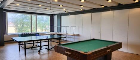 Uma sala de jogos com mesa de bilhar, mesa de futebol e piso de madeira, além de amplas janelas.