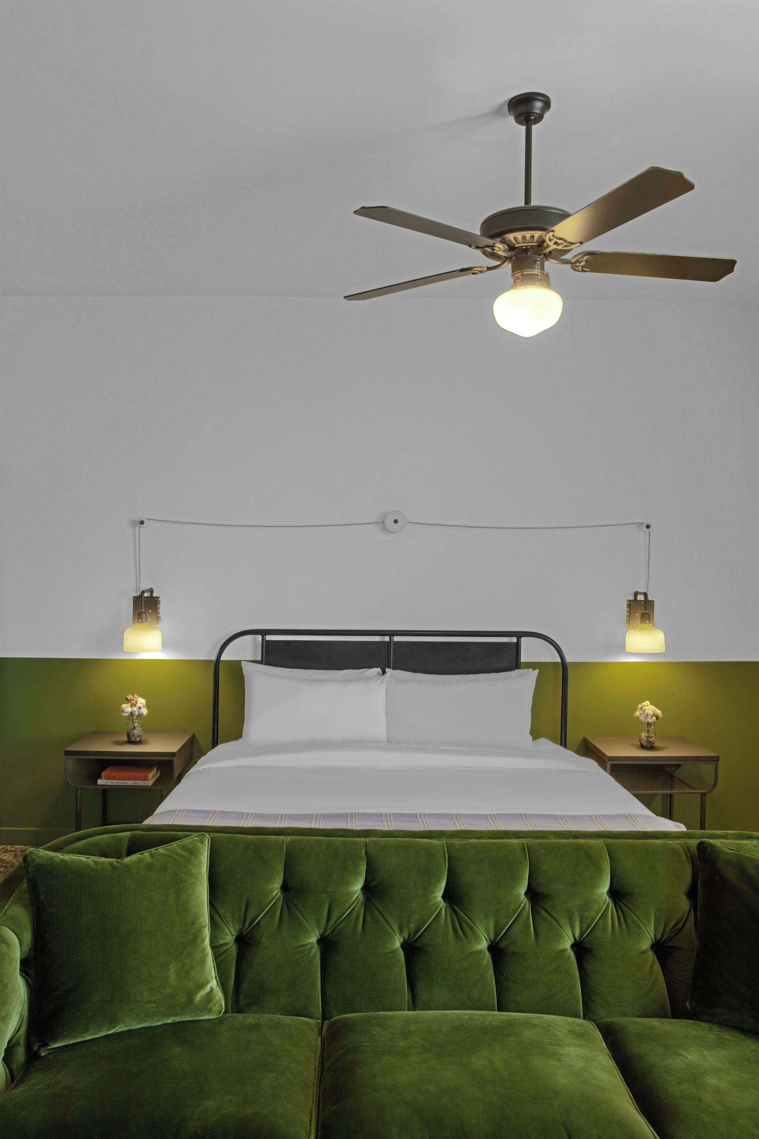 Quarto com cama, cabeceira estofada em verde, duas mesas de cabeceira com luminárias, um ventilador de teto e um aplique de parede.