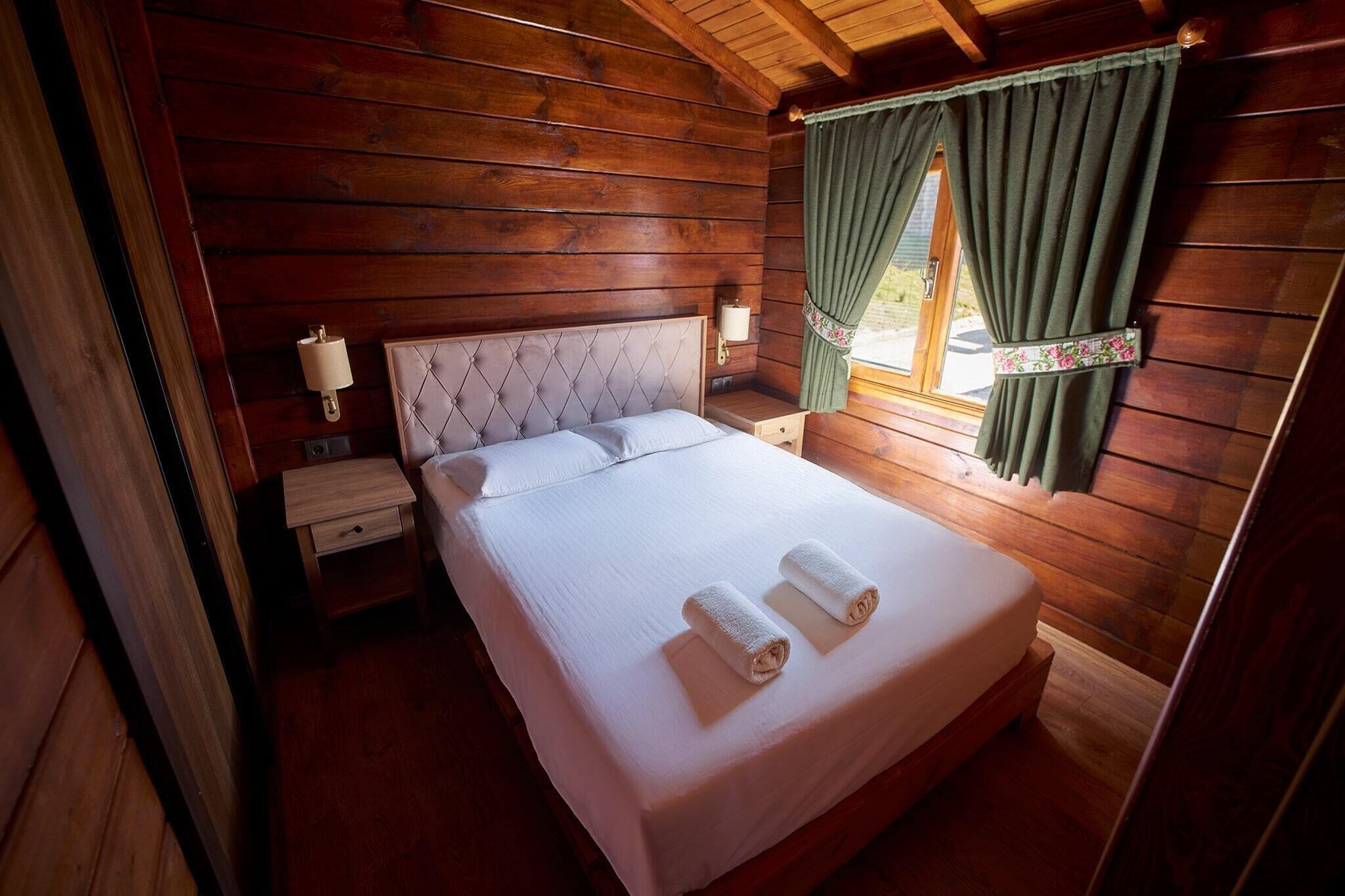 Quarto de cabana de madeira com cama, mesas de cabeceira e janela com cortinas.