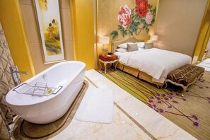 Quarto de hotel com uma banheira grande, cama com roupa de cama branca, mesa de cabeceira com luminárias e um quadro floral na parede.