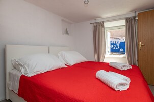 Uma cama bem arrumada com almofadas brancas e um edredom vermelho, uma toalha enrolada sobre a cama e uma janela com cortinas.