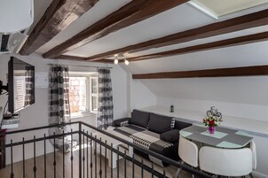 Quarto moderno no estilo loft com vigas de madeira aparentes, um sofá preto e branco, uma mesa de jantar e vista para a cidade pela janela.
