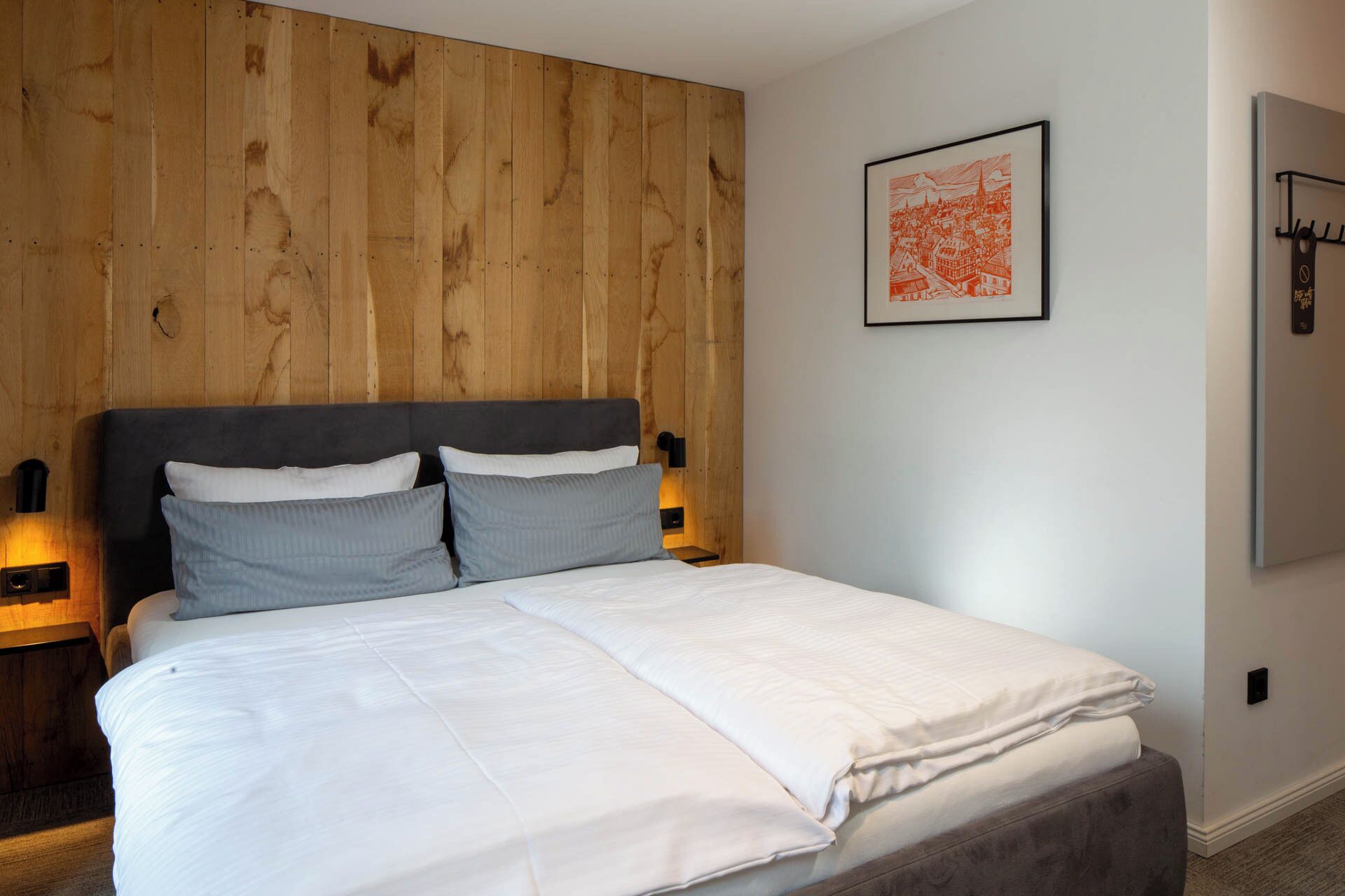 Cama bem arrumada com lençóis brancos e duas almofadas, cabeceira de madeira e uma obra de arte emoldurada na parede.