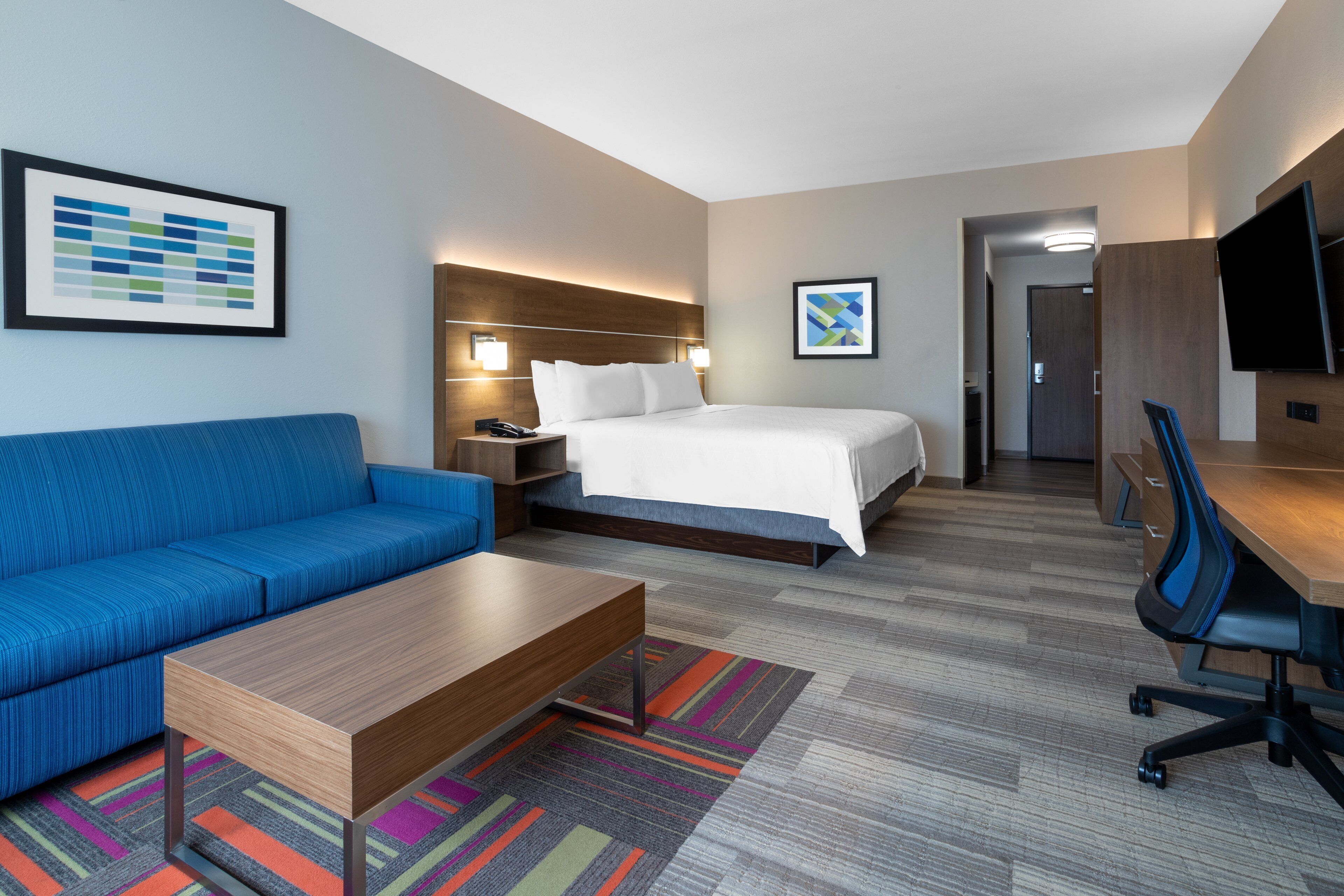 Quarto de hotel com sofá azul, mesa de centro de madeira, cama com roupa de cama branca, escrivaninha com cadeira e televisão na parede.