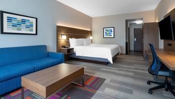 Quarto de hotel com sofá azul, mesa de centro de madeira, cama com roupa de cama branca, escrivaninha com cadeira e televisão na parede.