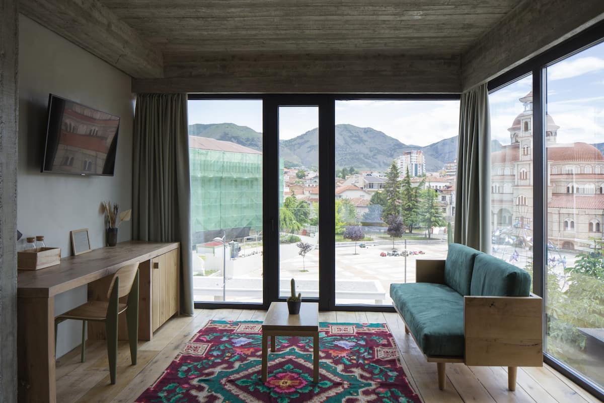 Uma sala de estar moderna com uma escrivaninha de madeira, um sofá verde e uma grande janela com vista para uma paisagem urbana e montanhas.