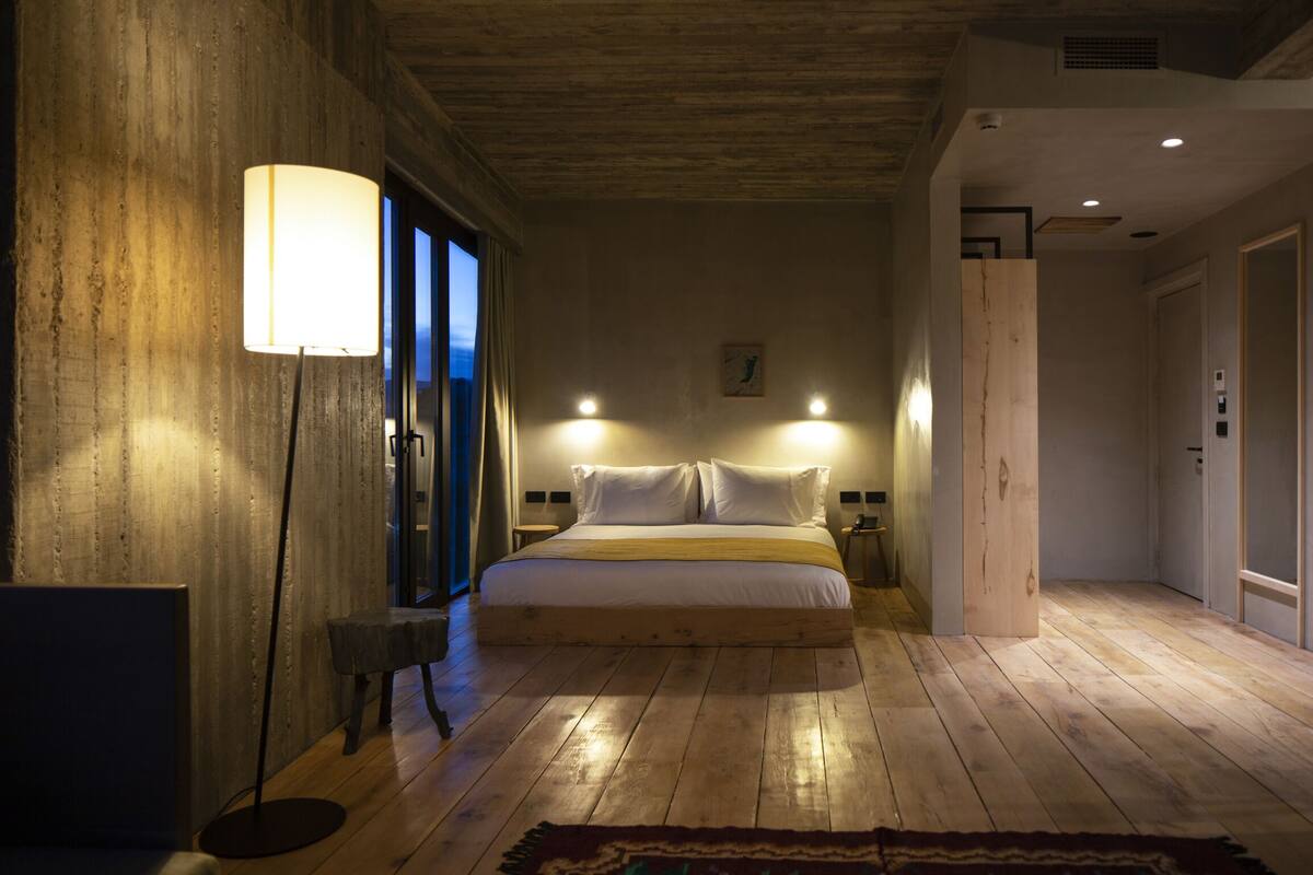 Quarto com piso de madeira, cama com roupa de cama branca, duas luminárias de cabeceira e vista pela janela.