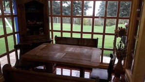 Uma mesa de jantar de madeira com bancos, um armário de madeira com diversos objetos e vista para um jardim gramado através de uma janela com padrão quadriculado.