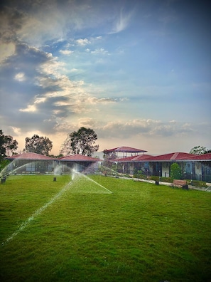 Um campo gramado com sistema de irrigação, um edifício com telhado vermelho e árvores ao fundo.