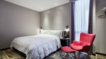 Quarto de hotel com cama, uma cadeira vermelha, mesa de cabeceira e janela com cortinas.