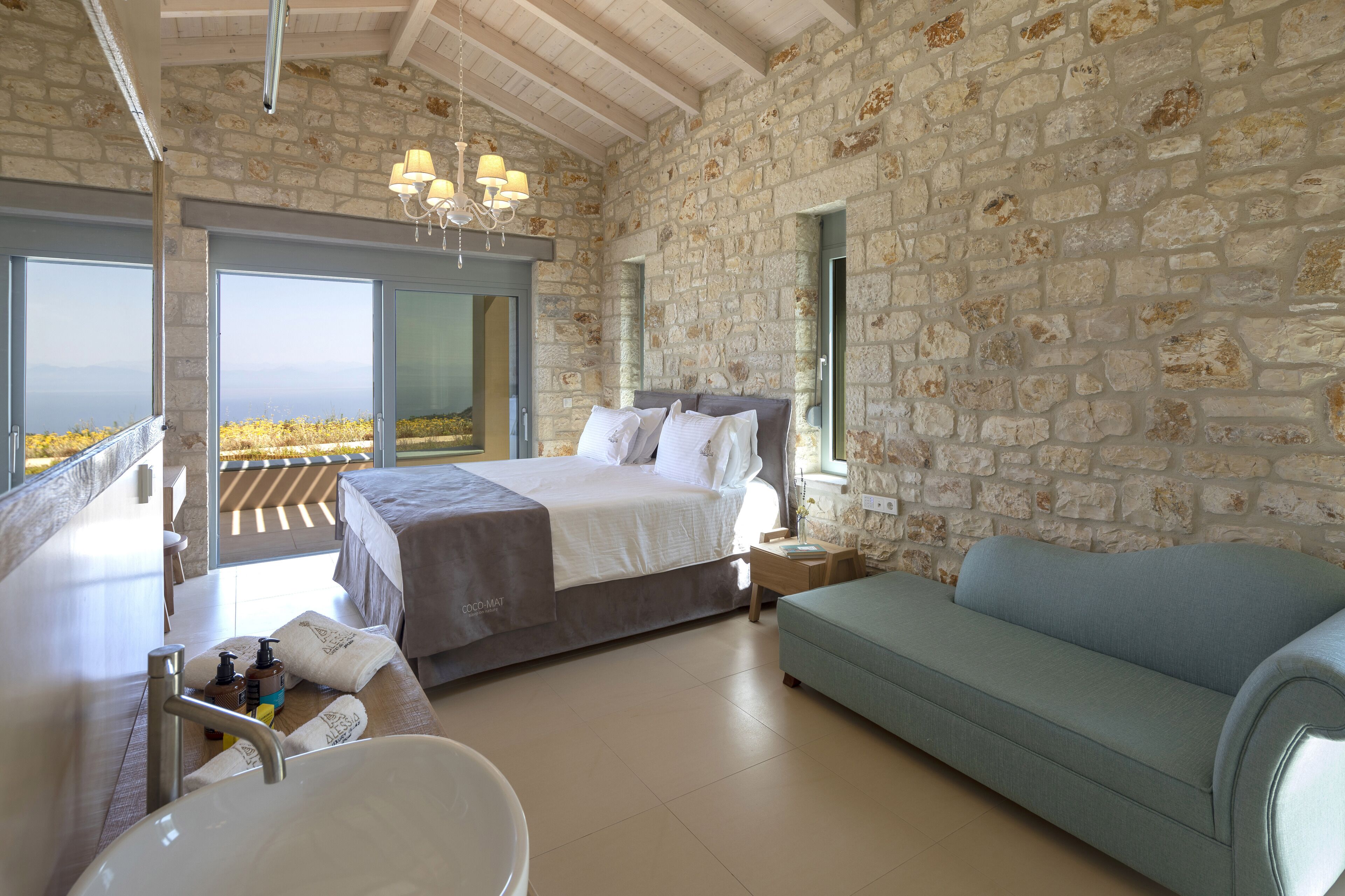 Quarto com parede de pedra, uma cama, um sofá, um candelabro e vista para a paisagem.