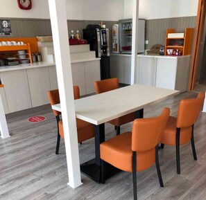 Uma área de refeições moderna com uma mesa branca e cadeiras laranja, uma máquina de café e uma geladeira ao fundo.
