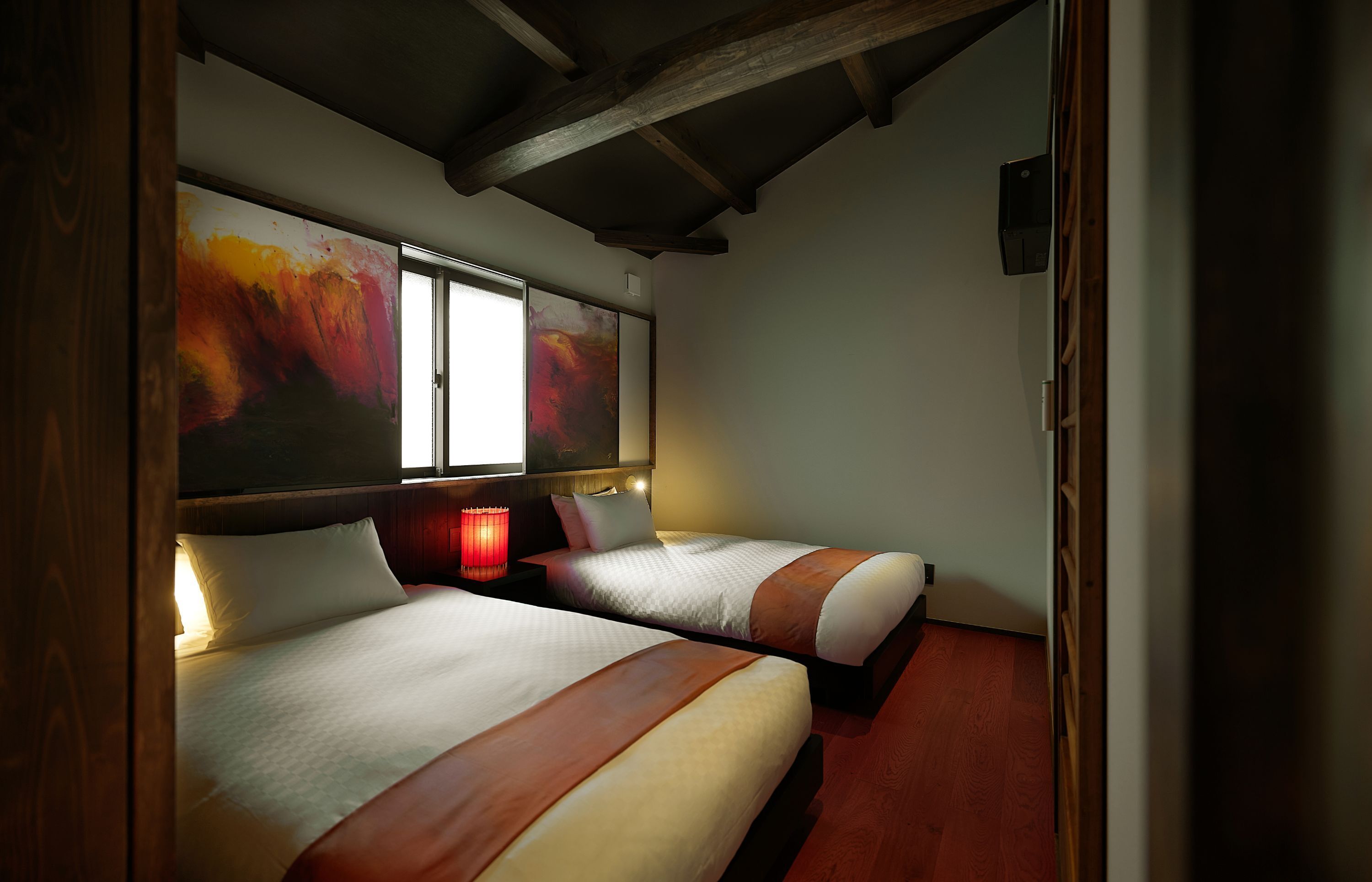 Quarto com duas camas, cabeceiras de madeira, um quadro abstrato grande e um abajur vermelho.