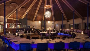 Ampla sala de jantar com mesas grandes preparadas para vários hóspedes, área de bar e teto alto com vigas de madeira.